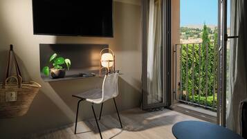 Quarto moderno com TV de tela plana, uma cadeira de vime, uma mesinha, abajur, planta e vista para o verde através de grandes janelas.