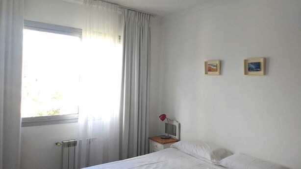 Um quarto com cama, mesa de cabeceira com abajur, janela com cortinas e duas imagens emolduradas na parede.