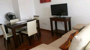 Uma sala de estar com uma mesa de jantar de madeira, cadeiras brancas, uma TV sobre um móvel, um sofá e um quadro com moldura vermelha na parede.