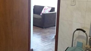 Um quarto com porta de madeira, piso de azulejos, sofá cinza com manta e cadeira azul com rodinhas.