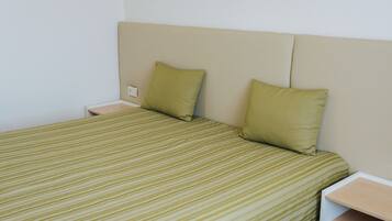Uma cama de solteiro com colcha verde listrada e duas almofadas, uma mesinha de cabeceira e um criado-mudo.