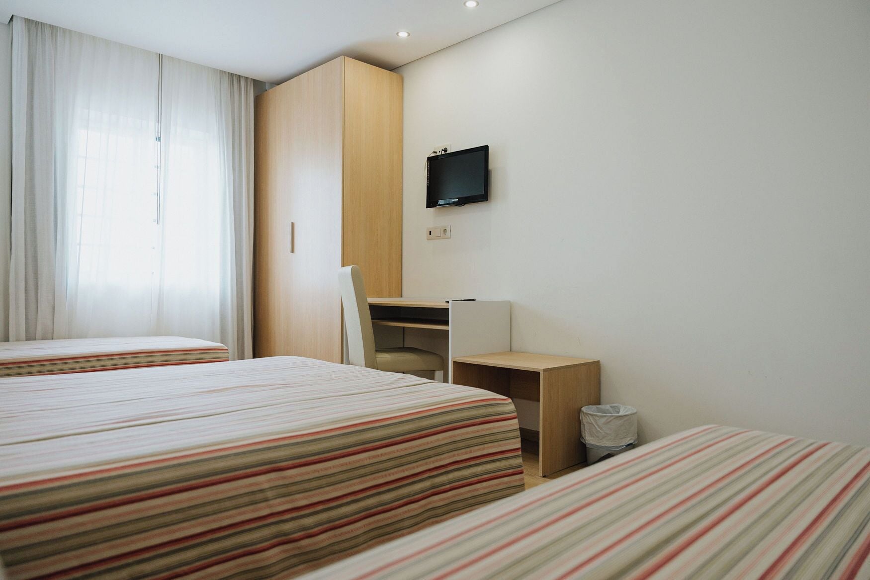 Quarto de hotel com duas camas, uma escrivaninha, uma cadeira, uma televisão e uma lixeira.