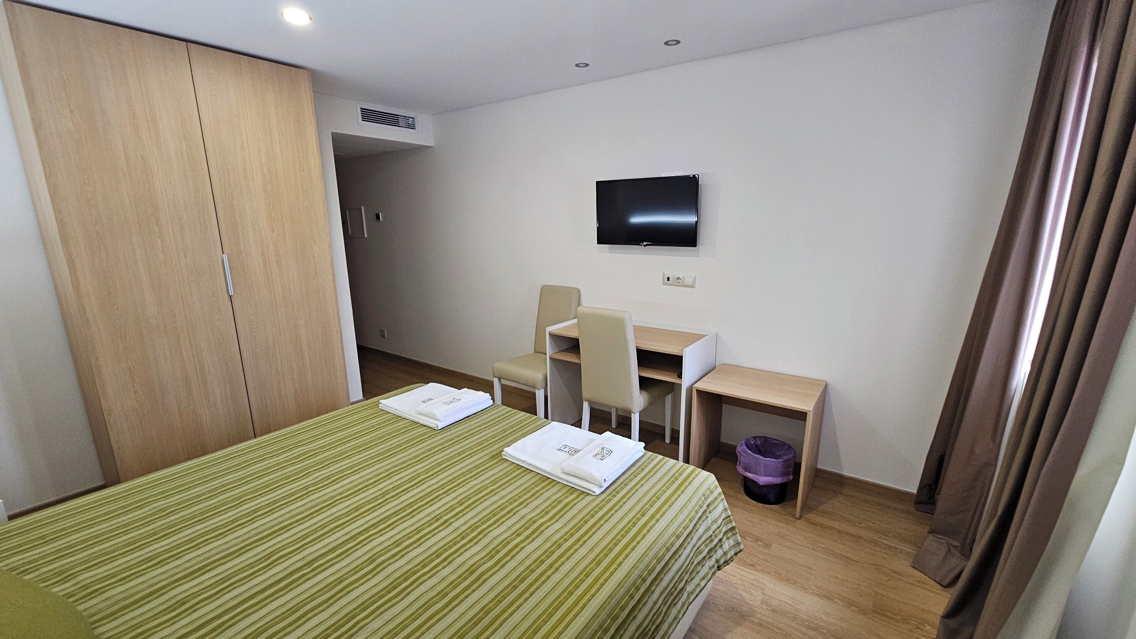 Quarto de hotel com cama, escrivaninha, cadeira, televisão e guarda-roupa.
