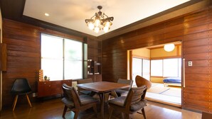 Uma sala de jantar com mesa e cadeiras de madeira, um candelabro e portas deslizantes que dão para um espaço com tatames.