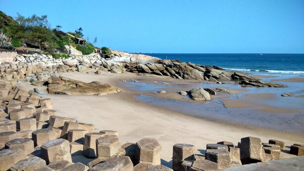 Uma praia com grandes blocos de concreto em primeiro plano, um litoral rochoso e um céu azul límpido.