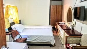 Quarto pequeno e compacto com uma cama de solteiro, cômoda de madeira, cadeira, escrivaninha e televisão.
