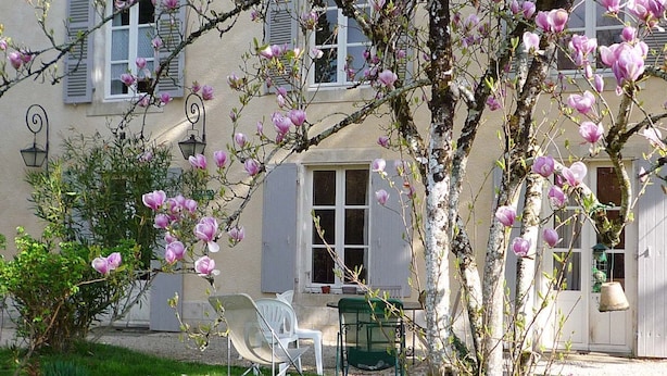 Um edifício com persianas brancas, um jardim com flores cor-de-rosa e mobiliário de exterior.
