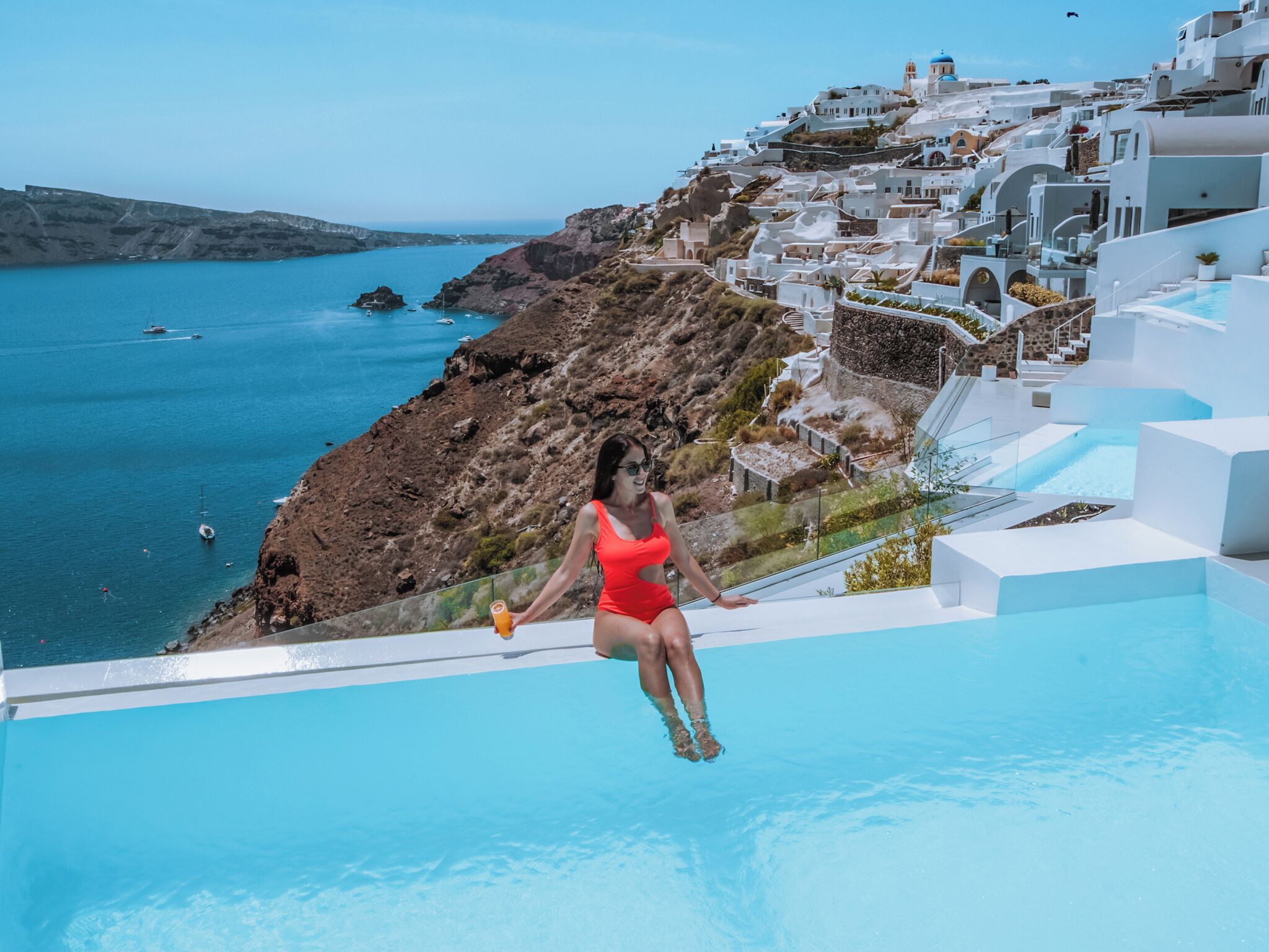 Mulher sentada na borda da piscina com vista para uma cidade costeira e o mar.