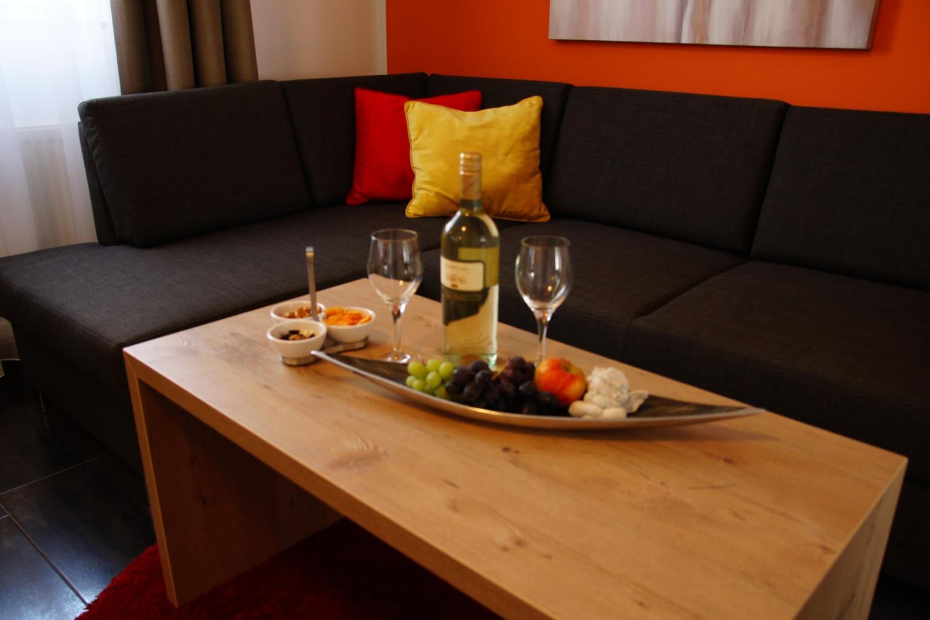 Uma mesa de jantar com um prato de frutas, uma garrafa de vinho e copos, colocada em frente a um sofá escuro com um travesseiro vermelho.