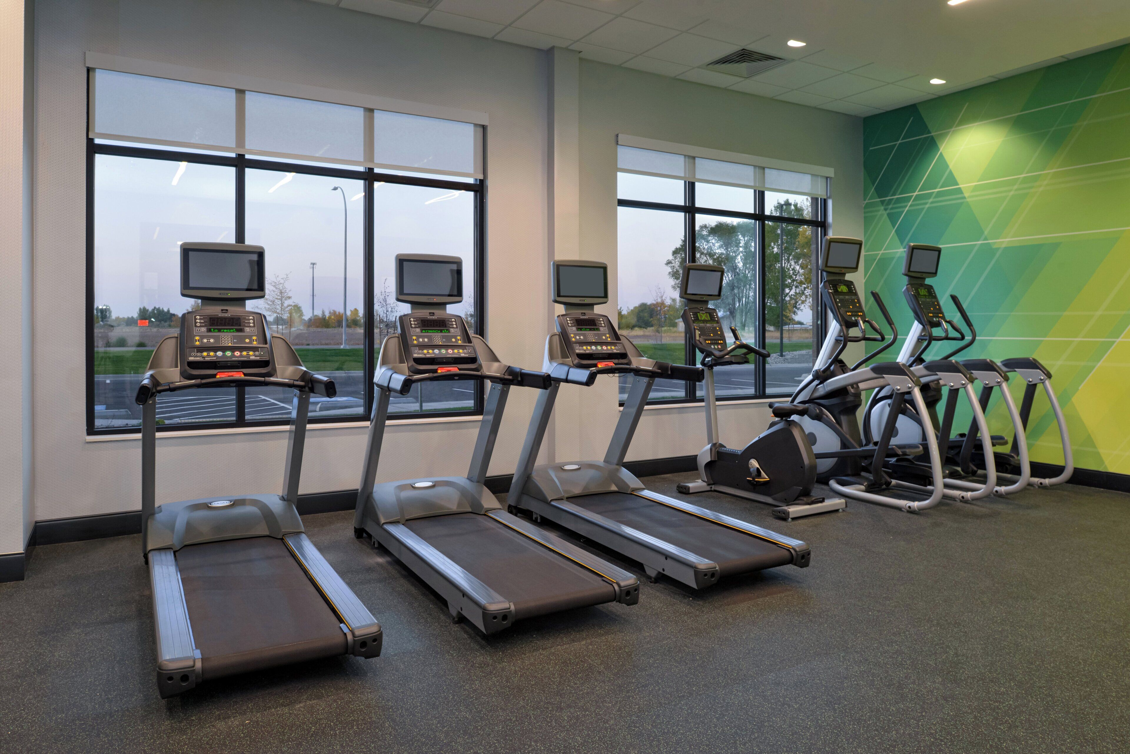 Academia com esteiras e bicicletas ergométricas, amplas janelas e uma parede verde.
