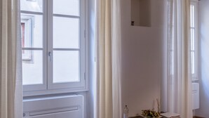 Quarto com uma janela grande, cortinas brancas, radiador, mesa de madeira com um vaso e um livro, e arcos de pedra no teto.