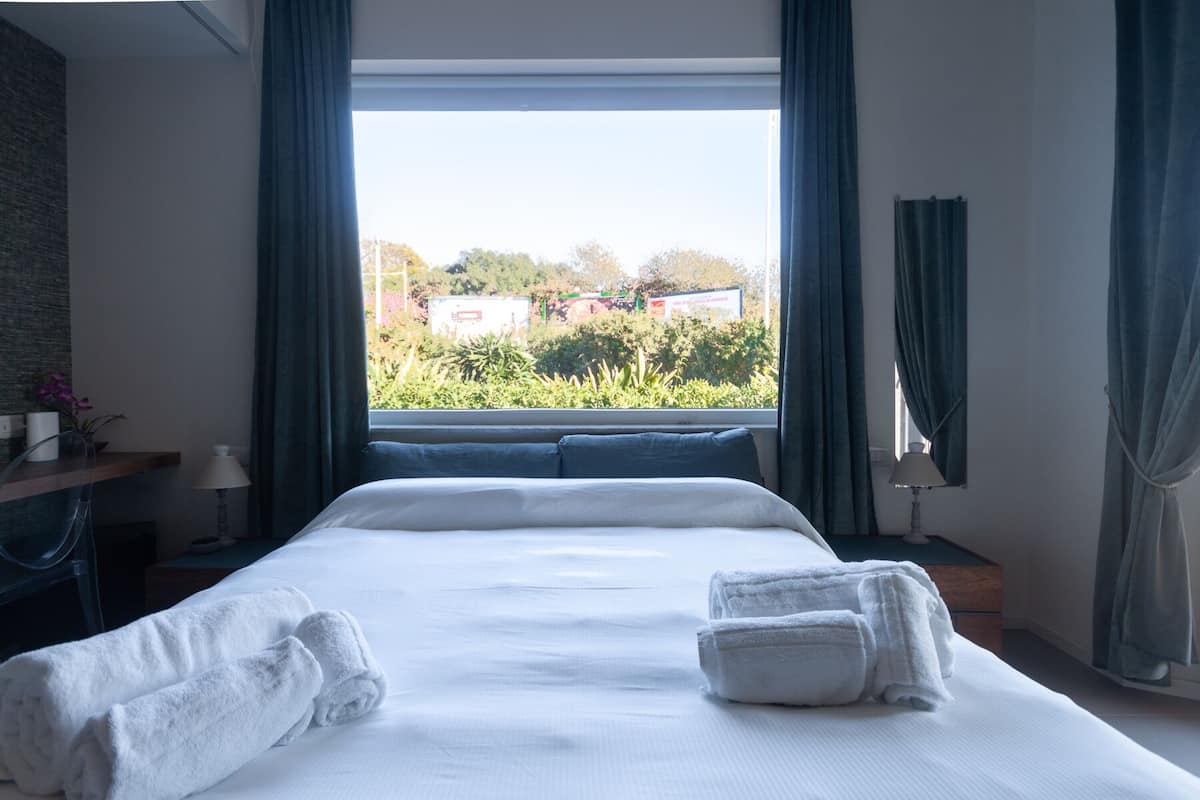 Quarto de hotel com uma cama grande, duas toalhas, uma mesa com abajur e vista para áreas verdes e edifícios.