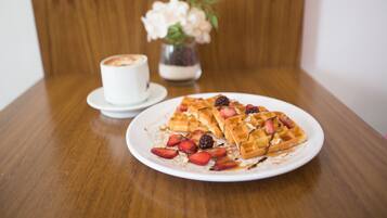 Uma pilha de waffles com morangos e framboesas, uma xícara de café e um vaso com flores sobre uma mesa de madeira.