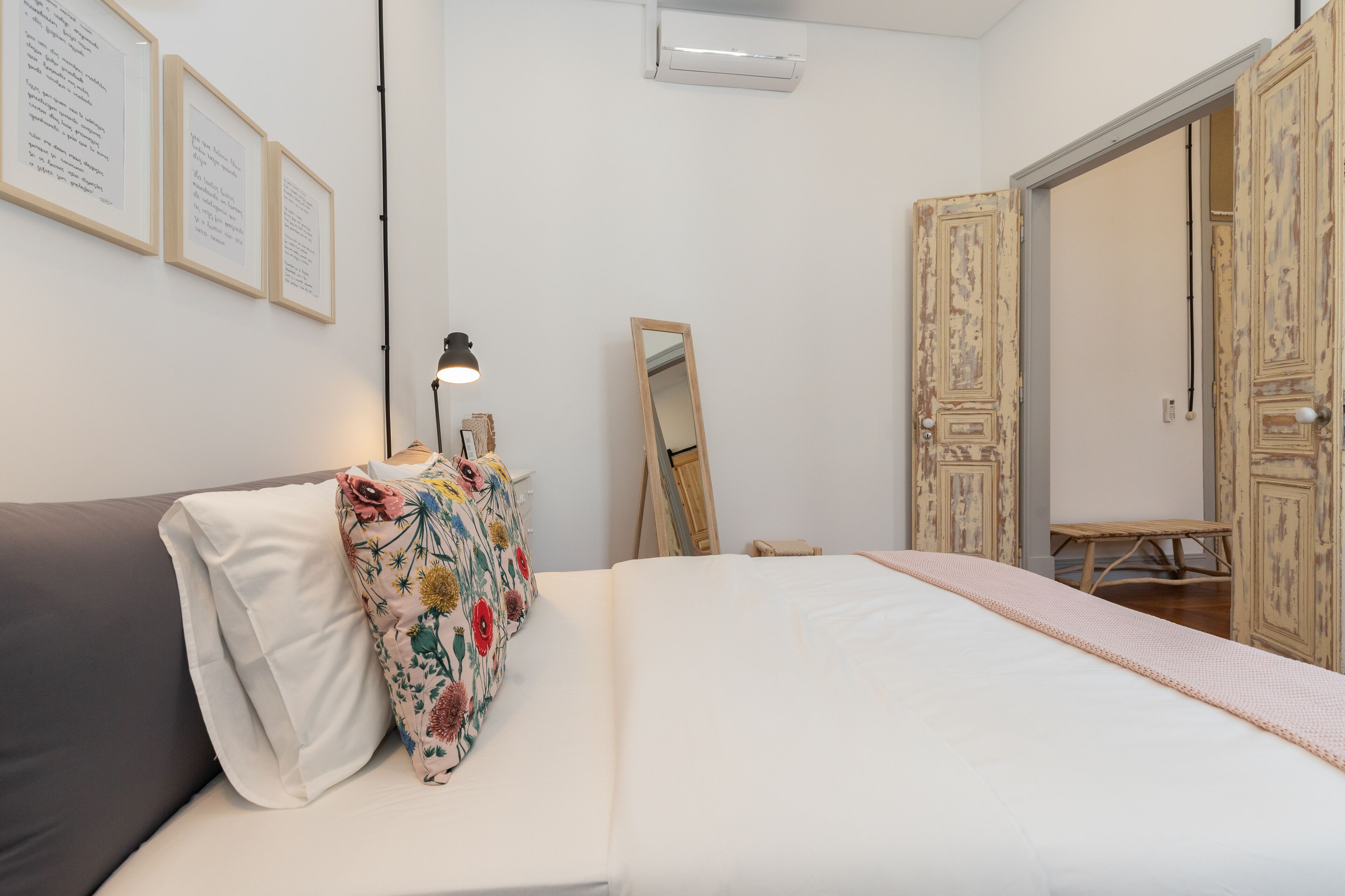 Quarto com cama, mesa de cabeceira com abajur, espelho e porta com moldura de madeira.