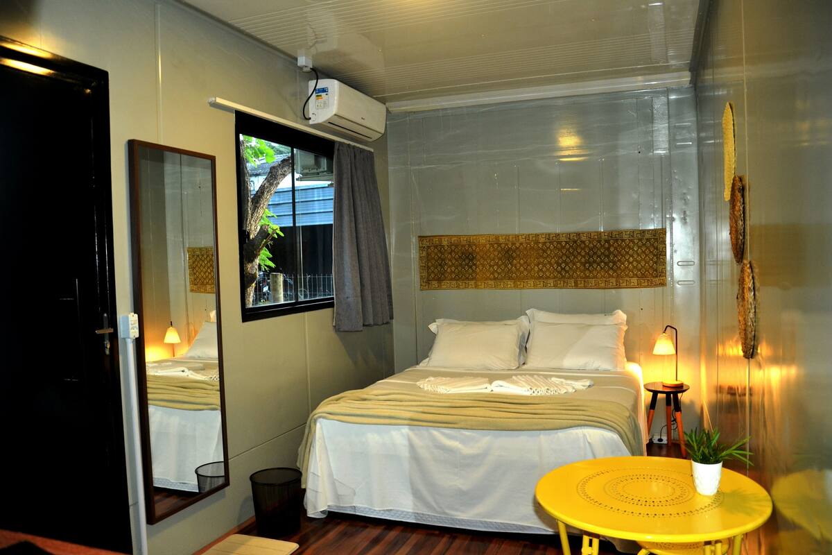 Quarto de hotel com cama, espelho, janela com cortinas, pequena mesa com planta e ar condicionado fixado na parede.