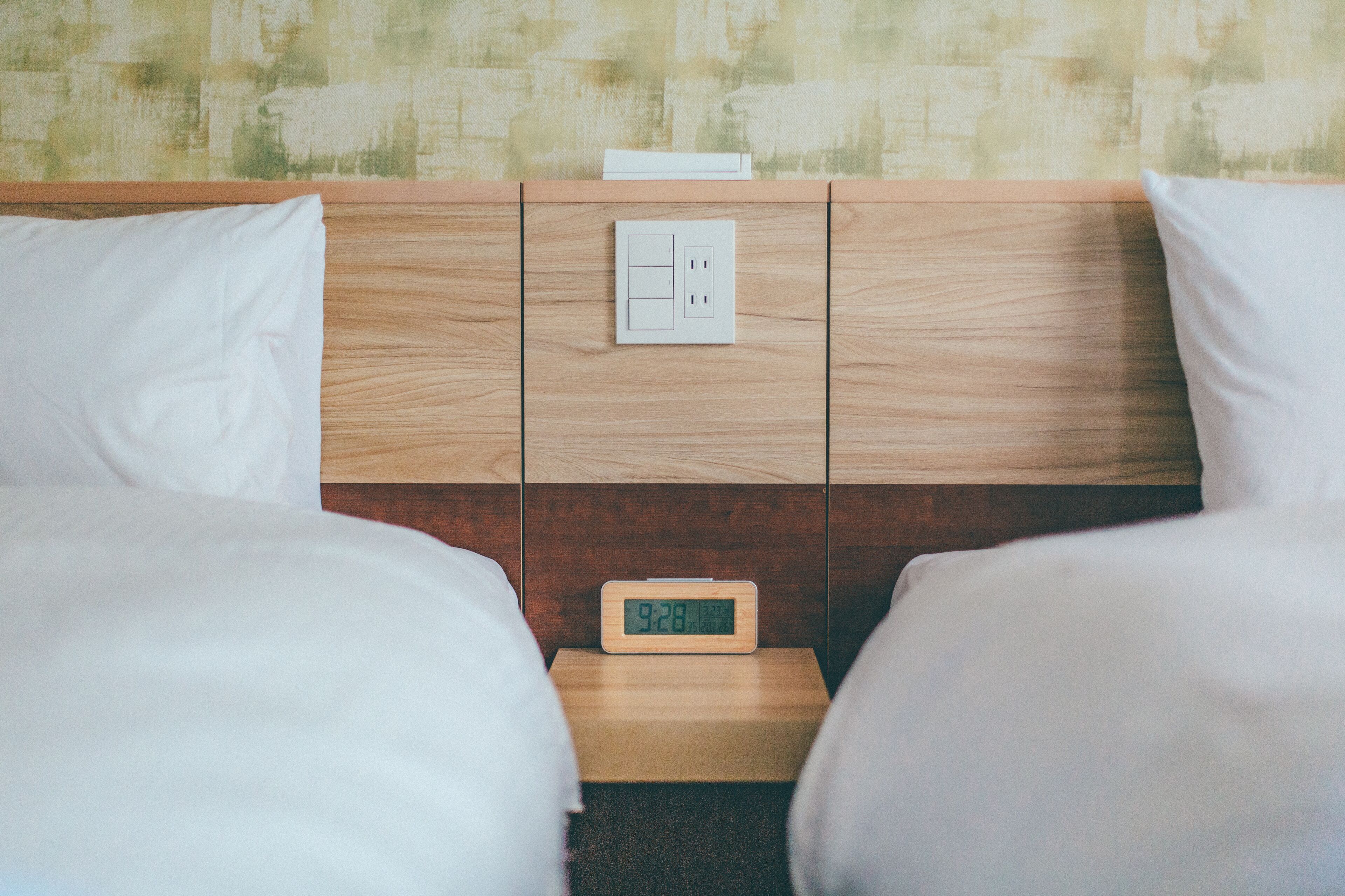 Quarto de hotel com duas camas, cabeceira de madeira, mesa de cabeceira com relógio digital e tomada.