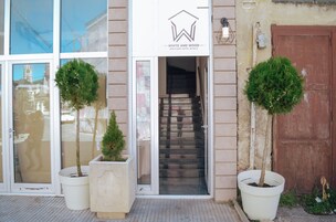 Entrada de um hotel com uma placa que diz 'WHITE AND WOOD BOUTIQUE HOTEL BITOLA'. Há duas árvores em vasos na frente da entrada.