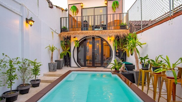 Uma área de piscina com um telhado de palha, plantas em vasos e uma escadaria que leva a uma varanda.