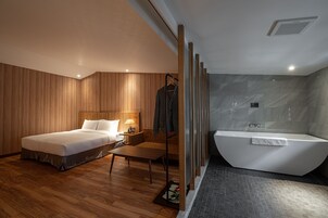 Quarto de hotel moderno com piso de madeira, uma cama grande, área de estar e banheiro separado com banheira.