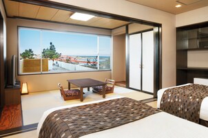 Quarto de hotel com uma grande janela que oferece vista para a área costeira, uma pequena mesa com cadeiras e duas camas com roupa de cama estampada.