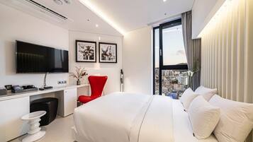 Quarto de hotel moderno com uma cama grande, uma escrivaninha com televisão de tela plana, uma cadeira vermelha e vista para a cidade pela janela.