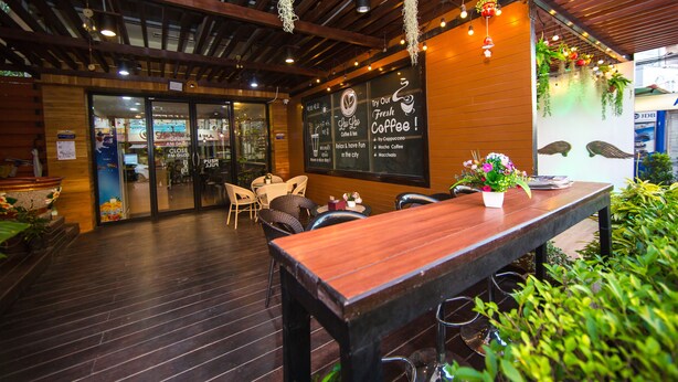 Uma cafeteria interna com piso de madeira, uma mesa comprida de madeira e cadeiras. Há luminárias pendentes e plantas.