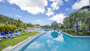 Piscina com cadeiras de descanso azuis, rodeada por gramado bem cuidado e palmeiras.