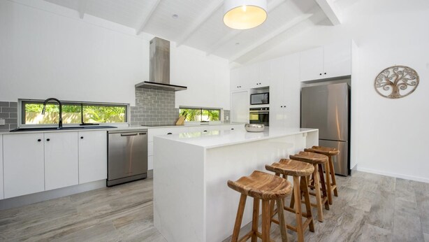 Cozinha moderna com armários brancos, uma ilha central com banquetas, eletrodomésticos de aço inoxidável e vista para o verde lá fora.