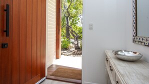 Uma porta de madeira dá acesso a uma área externa coberta, com uma árvore e uma parede branca com um espelho e uma tigela sobre um aparador.
