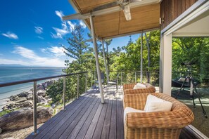 Um deck de madeira com um sofá e cadeiras de vime, com vista para a costa rochosa e o mar.