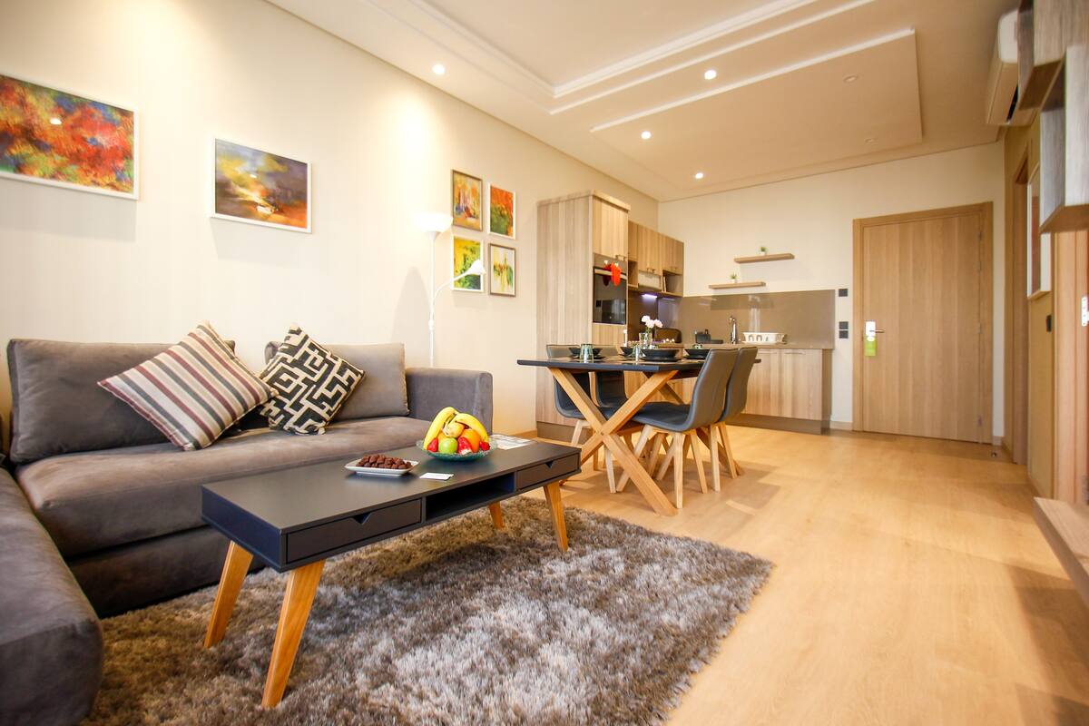 Uma sala de estar moderna com um sofá cinzento, uma mesa de centro preta e uma área de jantar com mesa e cadeiras de madeira.