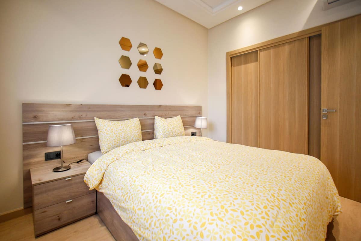 Quarto com cabeceira de madeira, duas mesas de cabeceira com luminárias, uma cama grande com colcha estampada e um guarda-roupa de madeira.