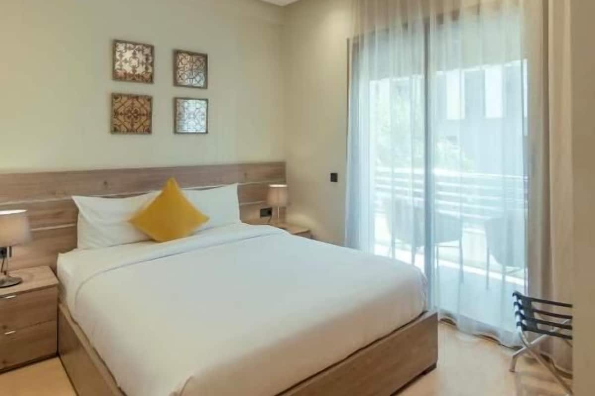 Quarto de hotel moderno com cama grande, cabeceira de madeira e vista do exterior através de cortinas leves.