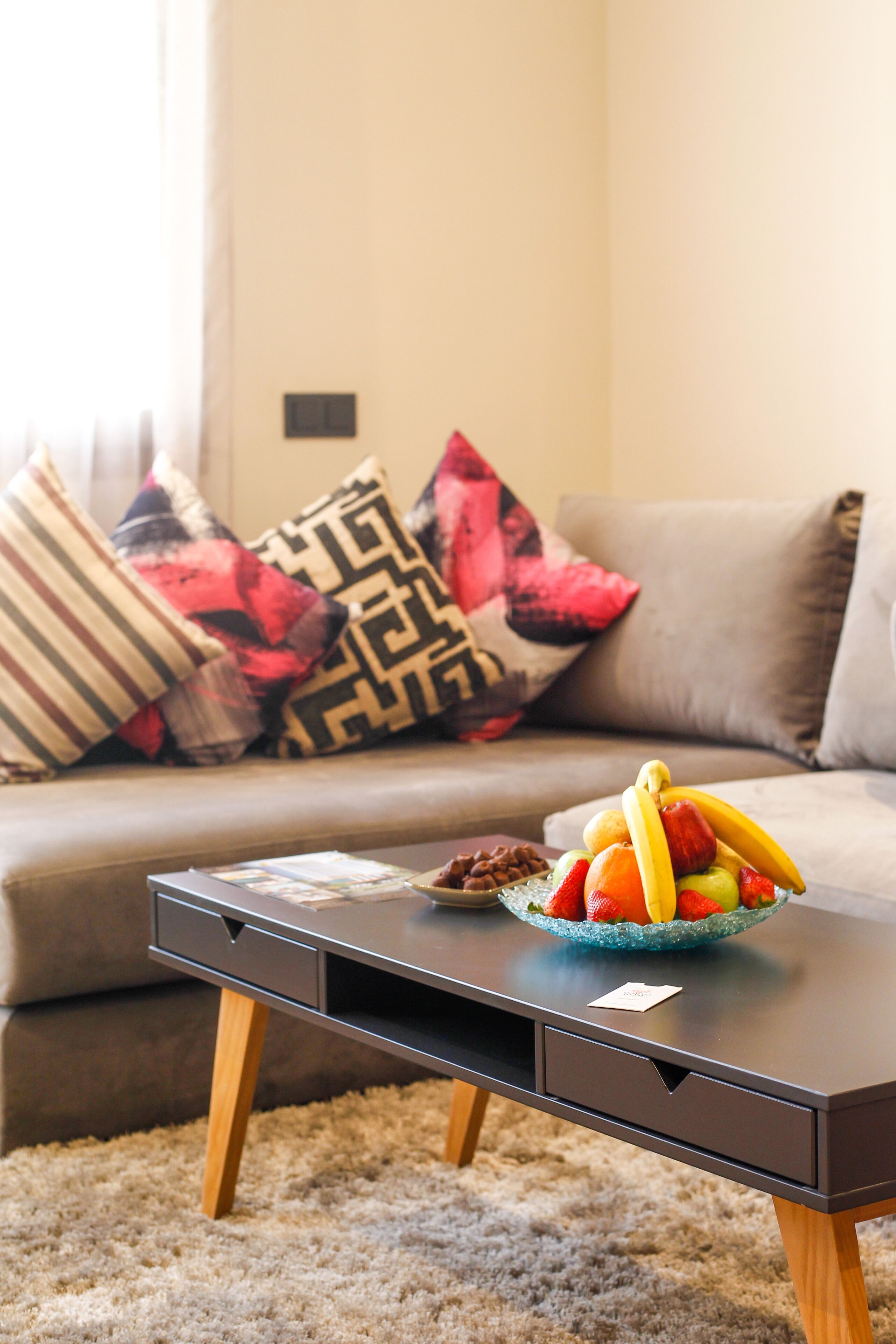 Uma sala de estar moderna com um sofá cinzento, almofadas estampadas, uma mesa de centro com uma fruteira e um pequeno objeto decorativo sobre a mesa.
