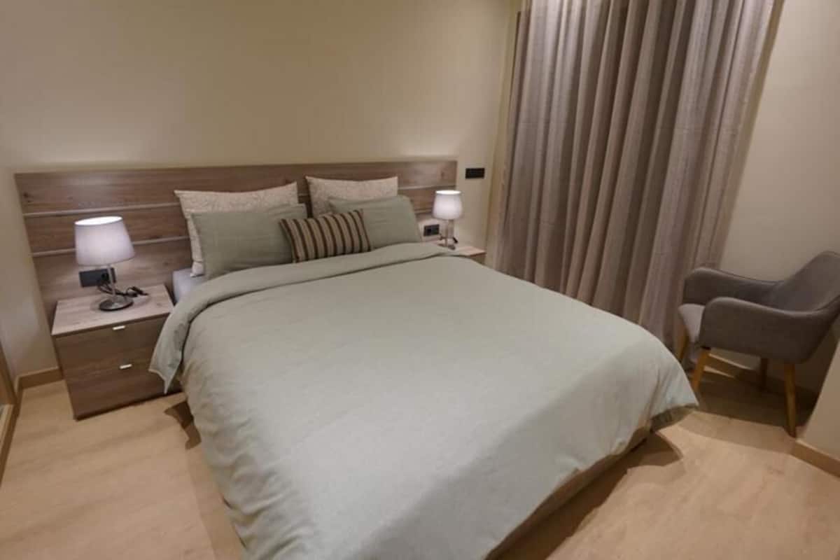 Uma cama bem arrumada com um cobreleito claro, duas mesas de cabeceira com abajures, uma cadeira e uma cortina.