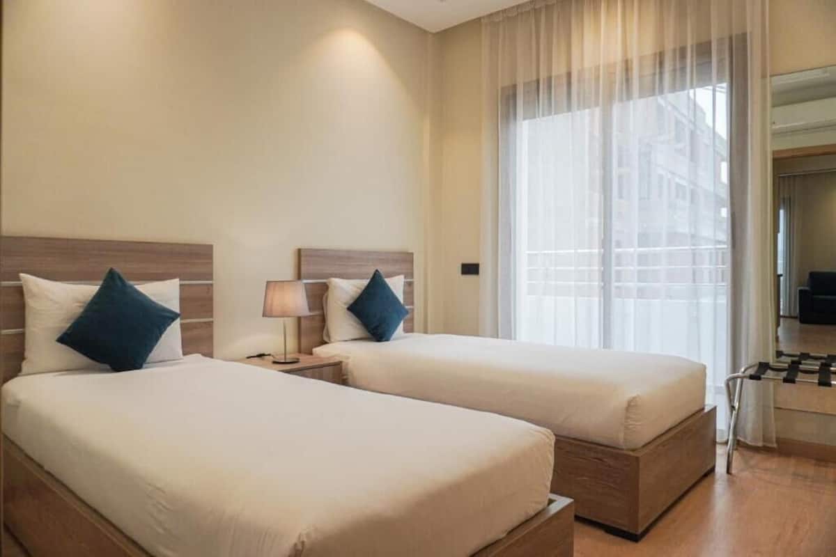 Quarto de hotel com duas camas, uma janela grande com cortinas finas e vista para a paisagem urbana.