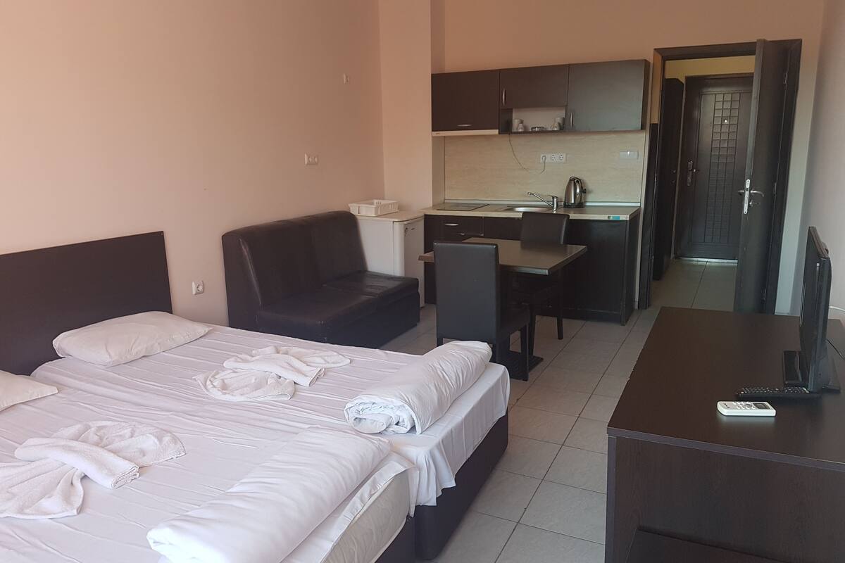 Quarto de hotel com duas camas, uma escrivaninha, uma cadeira, uma televisão e uma cozinha compacta.
