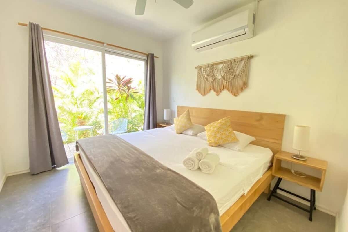 Quarto com uma cama grande, cabeceira de madeira, mesa de cabeceira com abajur e vista para o verde lá fora.