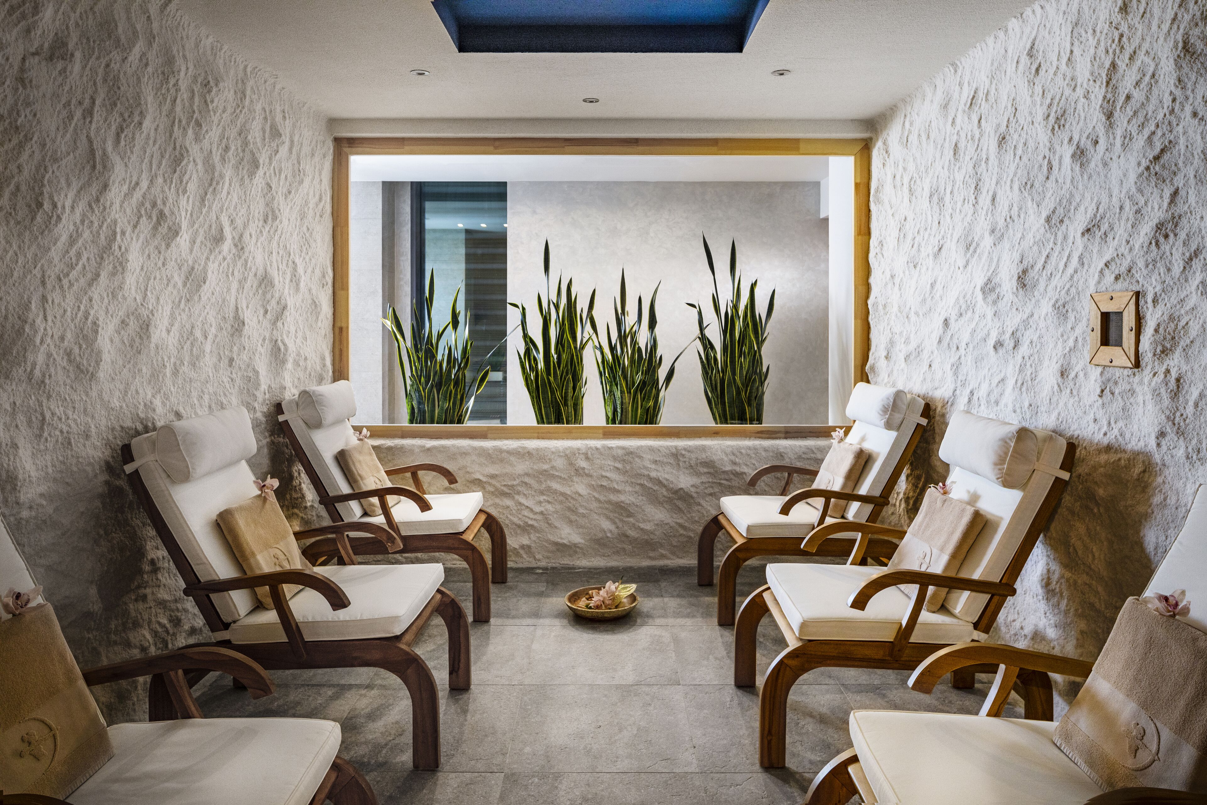 Um espaço de spa com seis cadeiras de madeira, uma janela grande com plantas e uma parede de pedra texturizada.