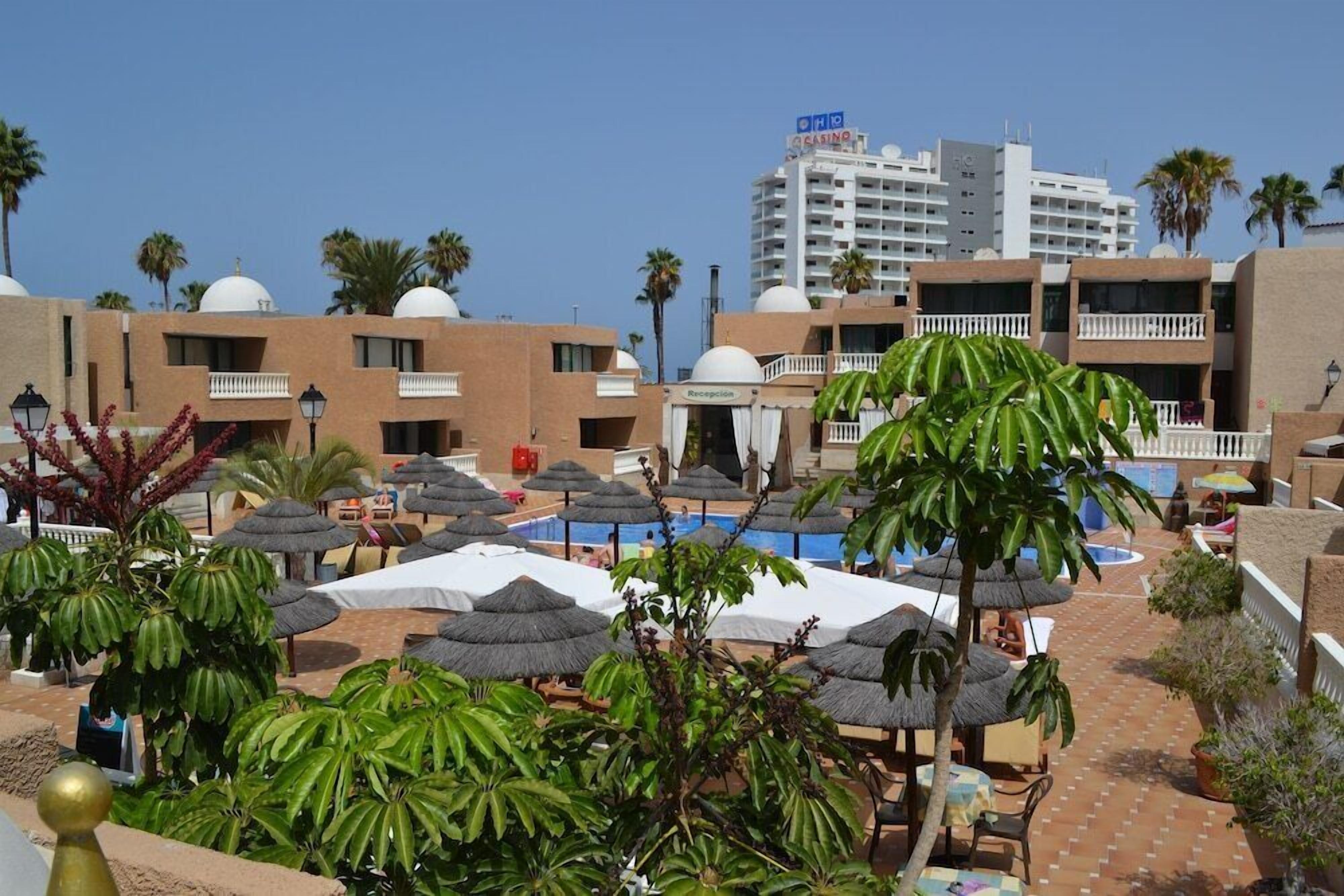 Resort com vários edifícios, área de piscina com guarda-sóis e palmeiras.