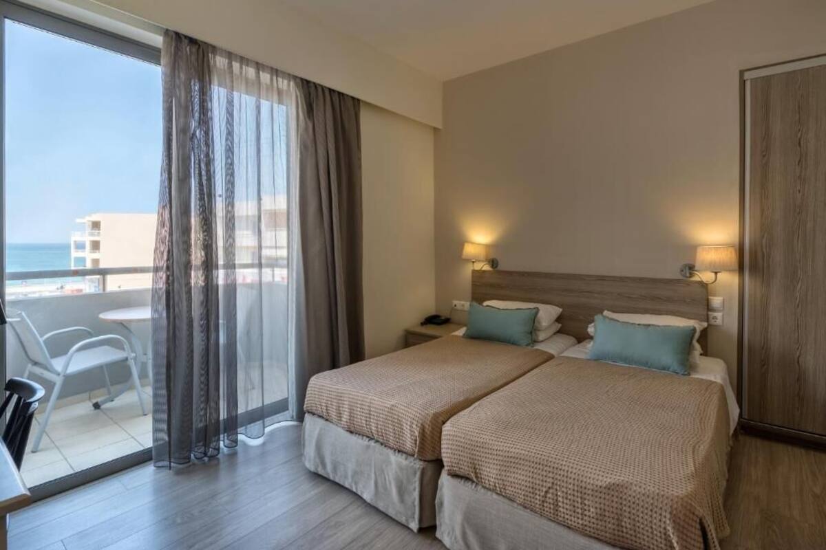 Quarto de hotel com duas camas, varanda com uma cadeira e vista para o mar.