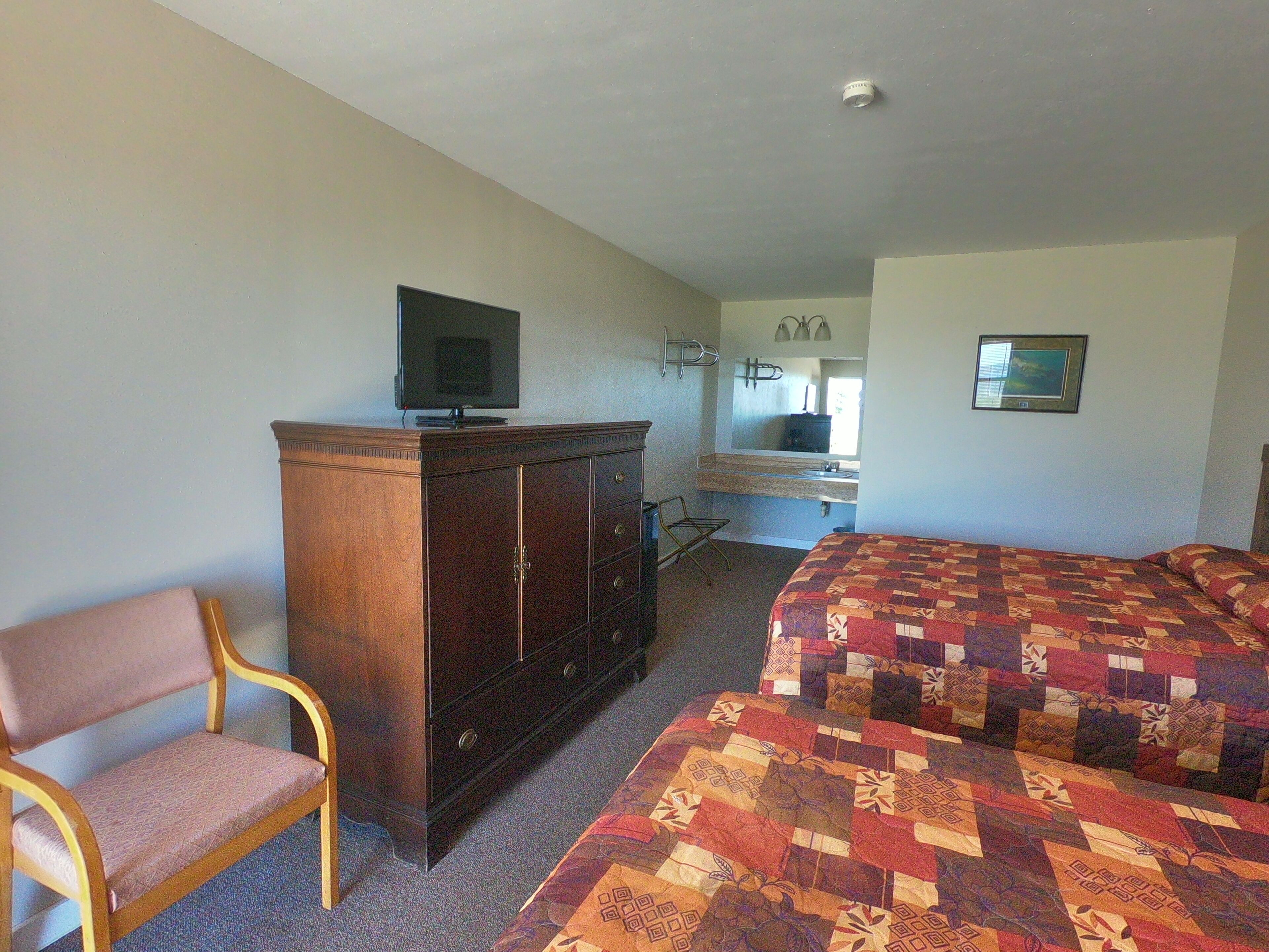 Quarto com cama, cômoda de madeira, televisão, cadeira e banheiro com pia e espelho.