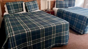 Quarto de hotel com duas camas, cada uma com roupa de cama xadrez combinando, cabeceira de madeira, mesa de cabeceira com abajur e porta com puxador dourado.