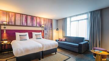 Quarto de hotel com uma cama grande, um sofá, uma mesinha e uma janela com cortinas.