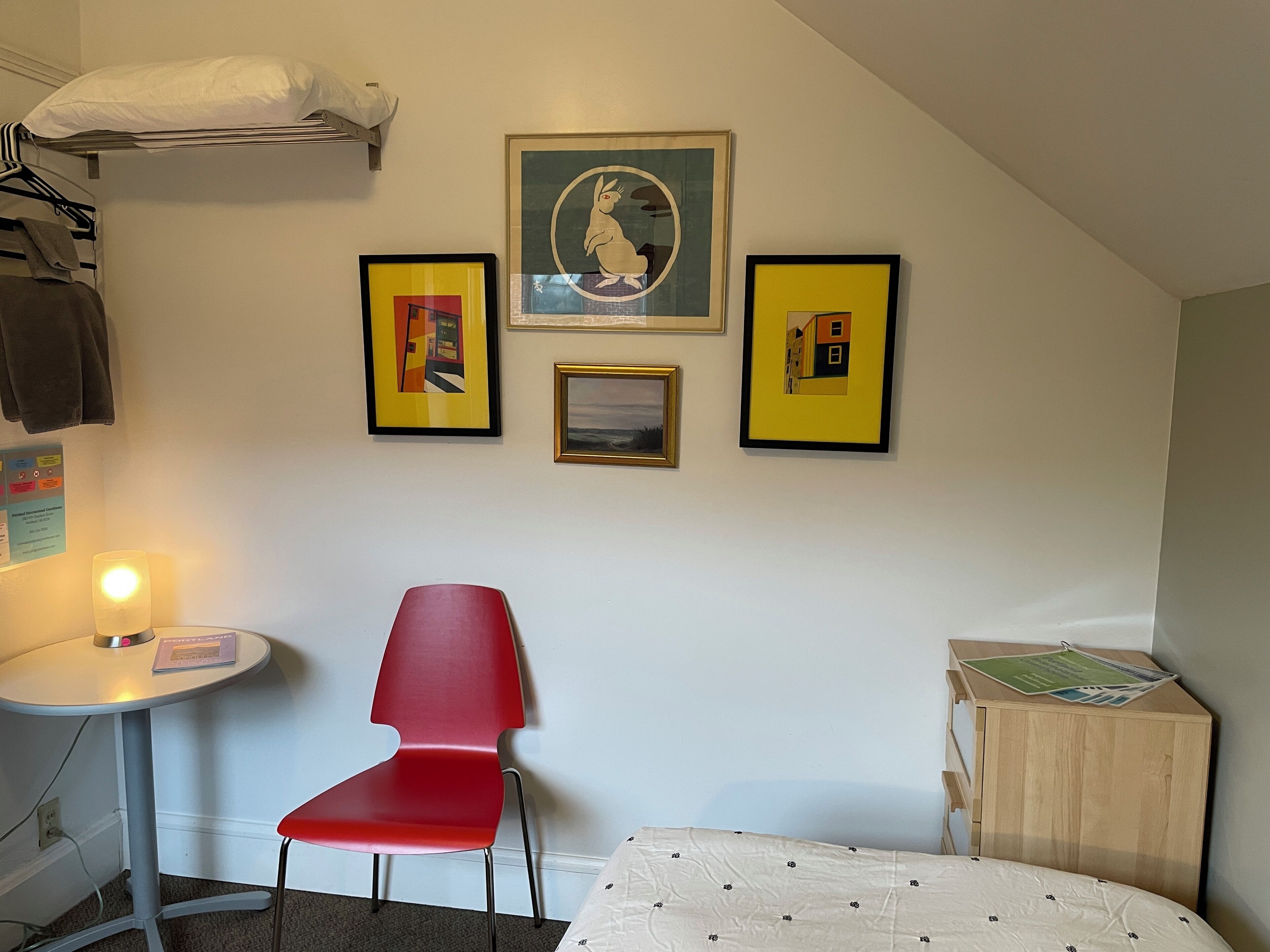 Um quarto com uma cama, uma cadeira vermelha, uma mesinha com abajur e três quadros na parede.