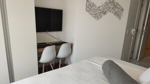 Quarto de hotel com cama, escrivaninha com duas cadeiras, televisão de tela plana e um objeto decorativo na parede.