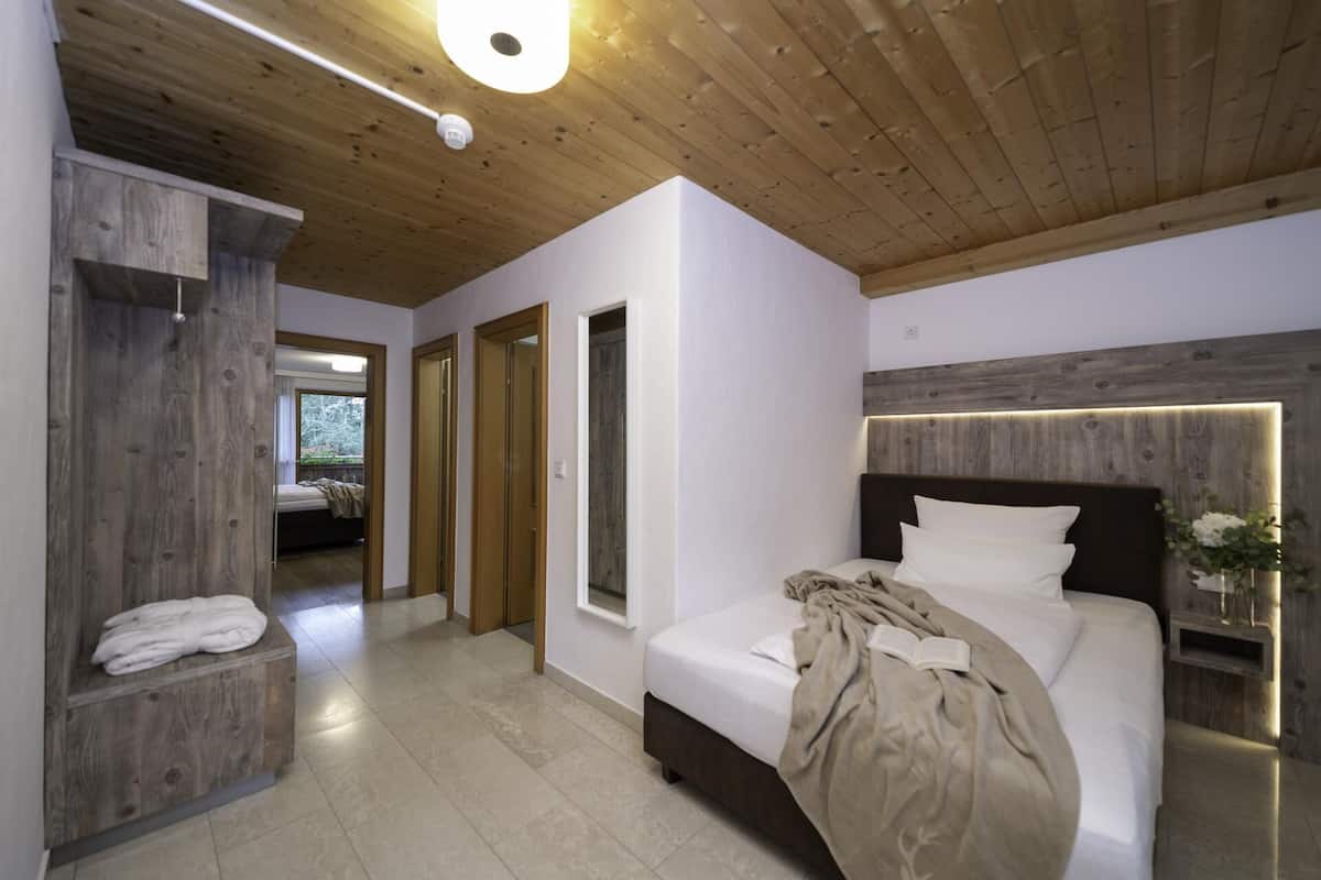 Quarto com teto de madeira, cama com roupa de cama branca, mesa de cabeceira com um vaso e vista para um jardim pela janela.