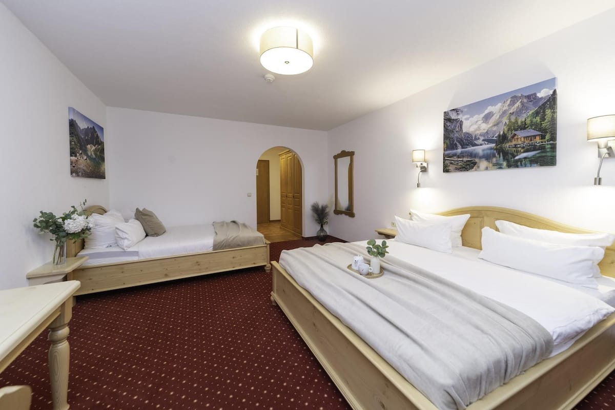 Quarto de hotel com duas camas de solteiro, cabeceira de madeira, tapete vermelho, uma mesinha, um vaso com flores e duas luminárias fixadas na parede.