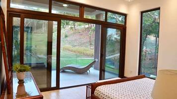 Quarto com cama, cómoda de madeira, ventoinha no teto e grandes portas de correr de vidro que dão para um jardim.