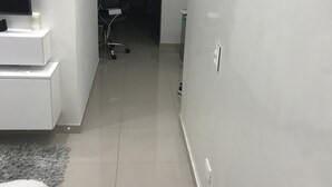 Corredor moderno de hotel com parede branca, TV de tela plana sobre um console branco, tapete cinza e piso de azulejos limpo.
